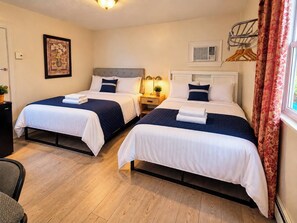 Quarto de hotel com duas camas, uma mesa de cabeceira com abajur, ar condicionado na parede e uma janela com cortinas.