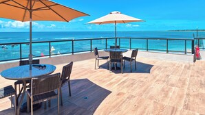 Um terraço com mesas e cadeiras redondas, dois grandes guarda-sóis e vista para o oceano.