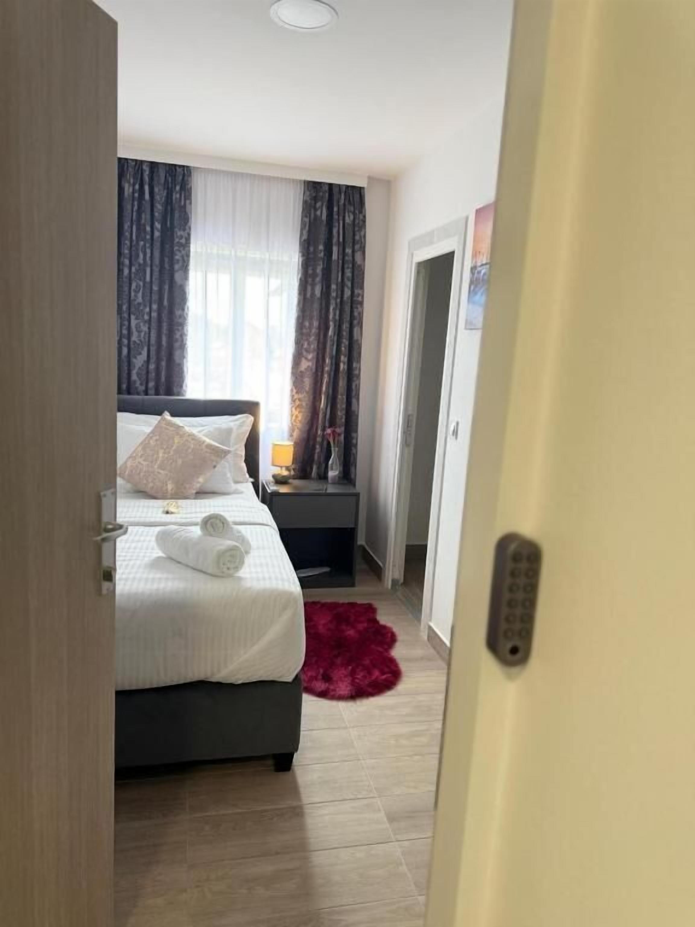Quarto de hotel com uma cama, uma mesa de cabeceira, um tapete vermelho e uma porta com fechadura eletrônica.