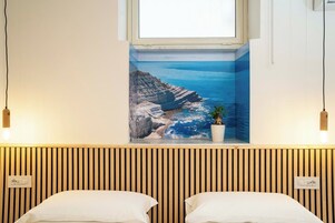 Quarto com parede de ripas de madeira, cama com roupa de cama branca, janela com mural costeiro e planta em vaso no parapeito.