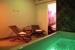 Um quarto de spa com duas poltronas de madeira, uma mesinha com um bule e uma banheira de hidromassagem verde.