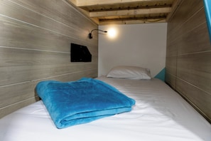 Quarto pequeno com cama de casal, paredes de madeira, colcha branca e manta azul.