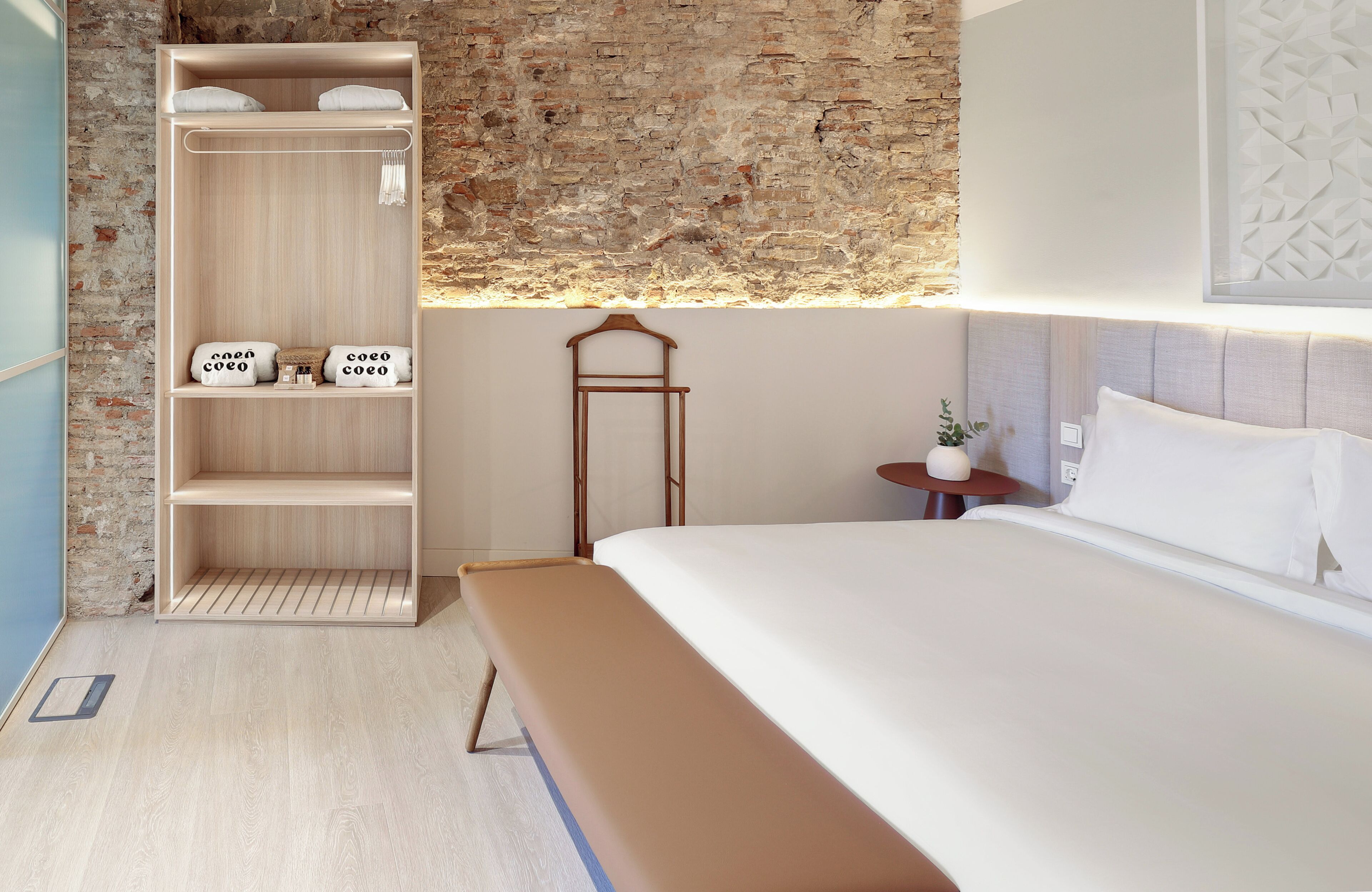 Quarto de hotel moderno com armário de madeira, cama com roupa de cama branca, parede de tijolos e uma pequena mesa de cabeceira com uma planta.