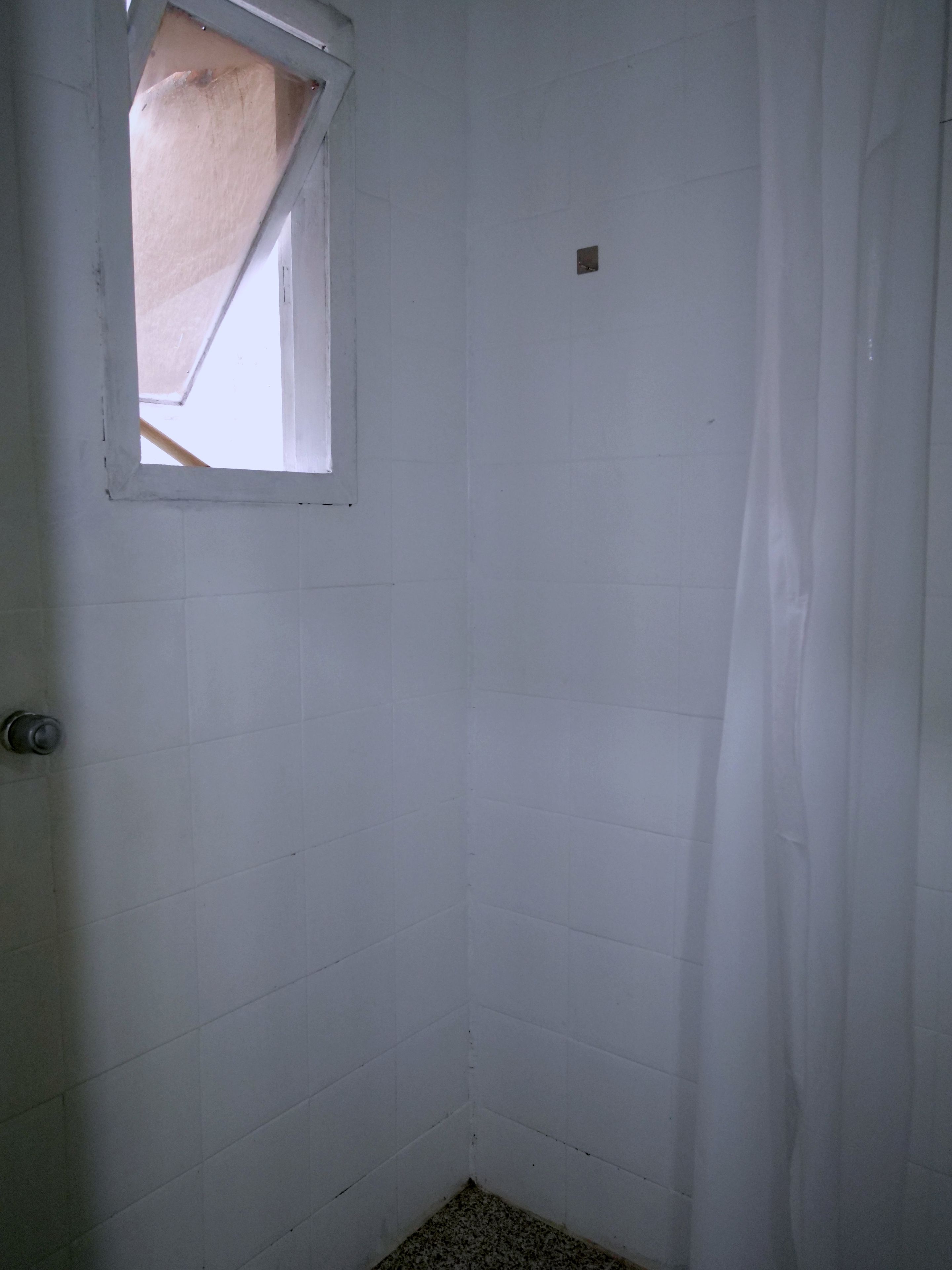 Um banheiro pequeno e fechado, com paredes em azulejo branco, uma janela e um cortina de chuveiro.