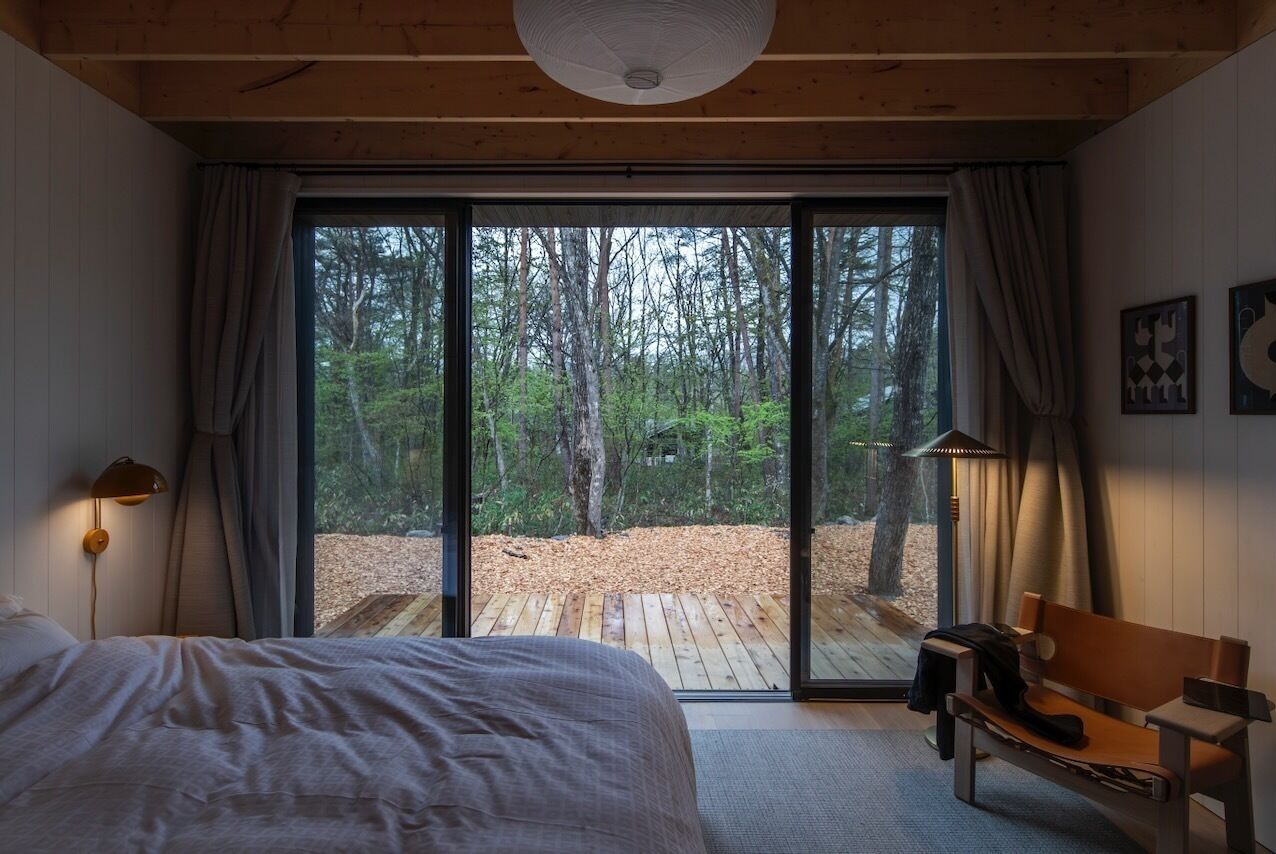 Quarto com uma grande porta de vidro deslizante que dá para um deck, uma cama, um banco e um abajur.