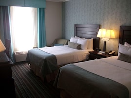 Quarto de hotel com duas camas, mesa de cabeceira com abajur, telefone e janela com cortinas.