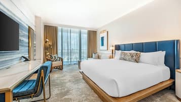 Quarto de hotel moderno com uma cama grande, uma escrivaninha, uma cadeira, uma televisão e uma janela ampla com cortinas.