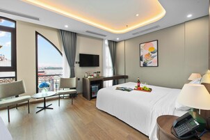Quarto de hotel com uma cama grande, uma escrivaninha, uma cadeira, uma mesinha de centro com um vaso de flores, uma televisão fixada na parede e uma varanda com vista.