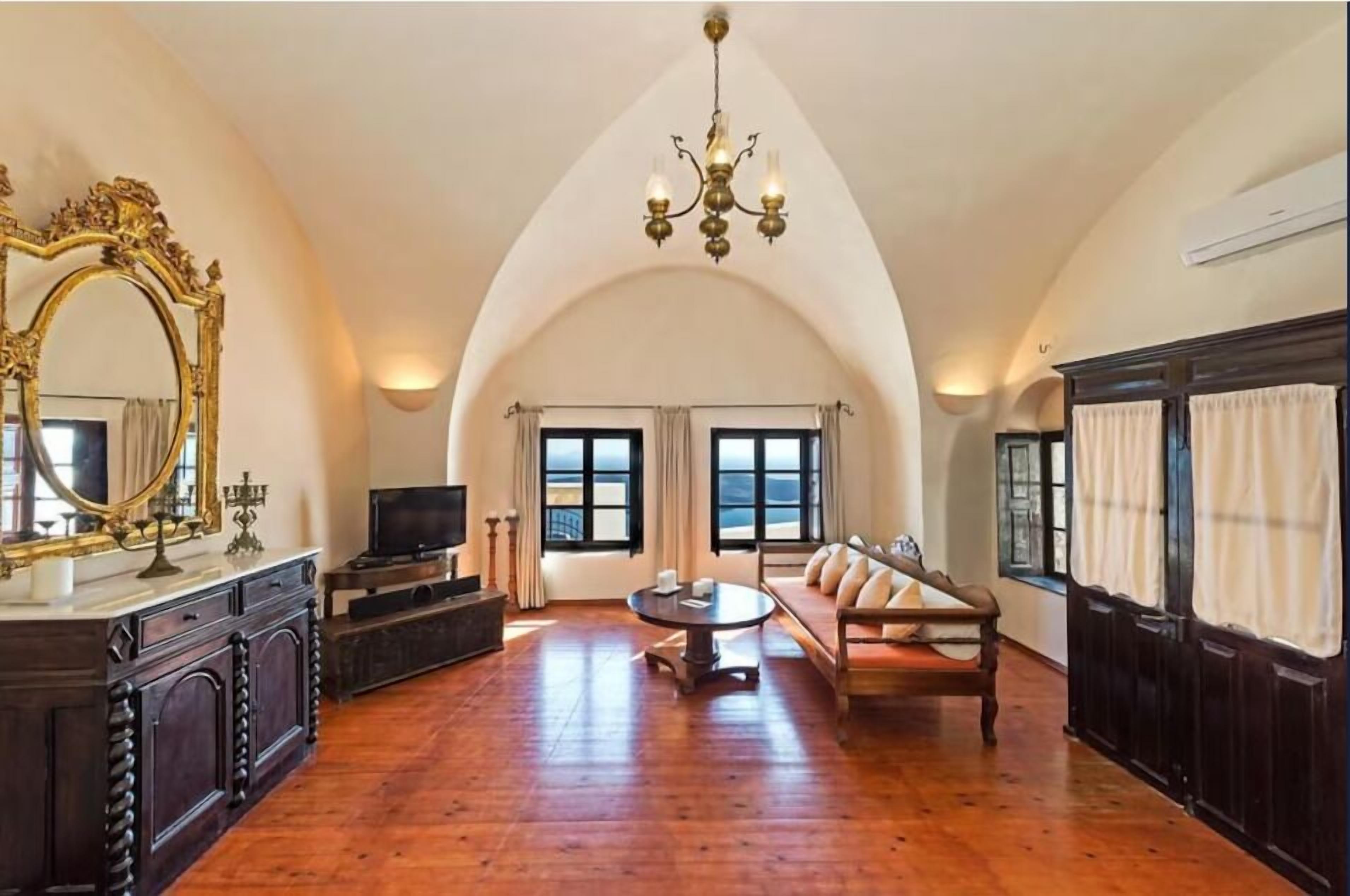 Um quarto espaçoso com teto alto em arco, um candelabro, um espelho grande, um sofá, uma televisão e vista para o mar através de amplas janelas.