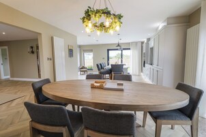Uma sala de jantar espaçosa com uma mesa redonda de madeira, rodeada por seis cadeiras, uma luminária pendente e vista para o pátio externo através de uma grande janela.