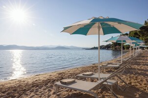 Uma praia com espreguiçadeiras e guarda-sóis, mar calmo e montanhas ao longe.