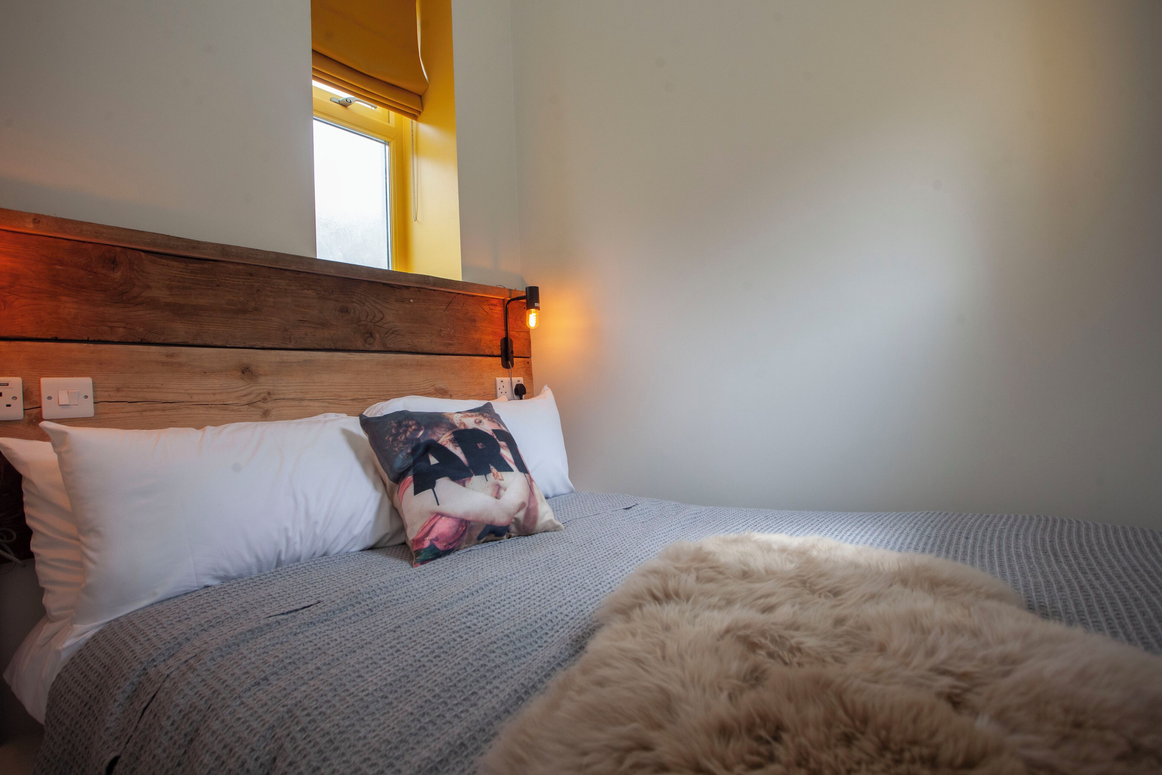 Quarto com cabeceira de madeira, cama com colcha estampada, tapete felpudo e luminária fixada na parede.