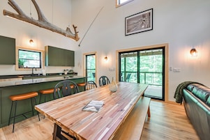 Cozinha moderna com mesa de madeira, banquetas de bar e um banco. Há duas janelas com vista para árvores.