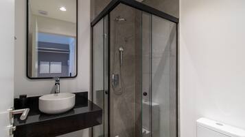Uma casa de banho moderna com chuveiro em cabine de vidro, lavatório branco e bancada escura.