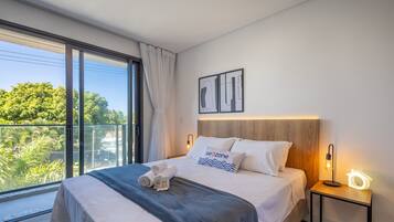 Quarto de hotel moderno com uma janela grande, cama com roupa de cama branca, cabeceira de madeira e mesa de cabeceira com abajur.