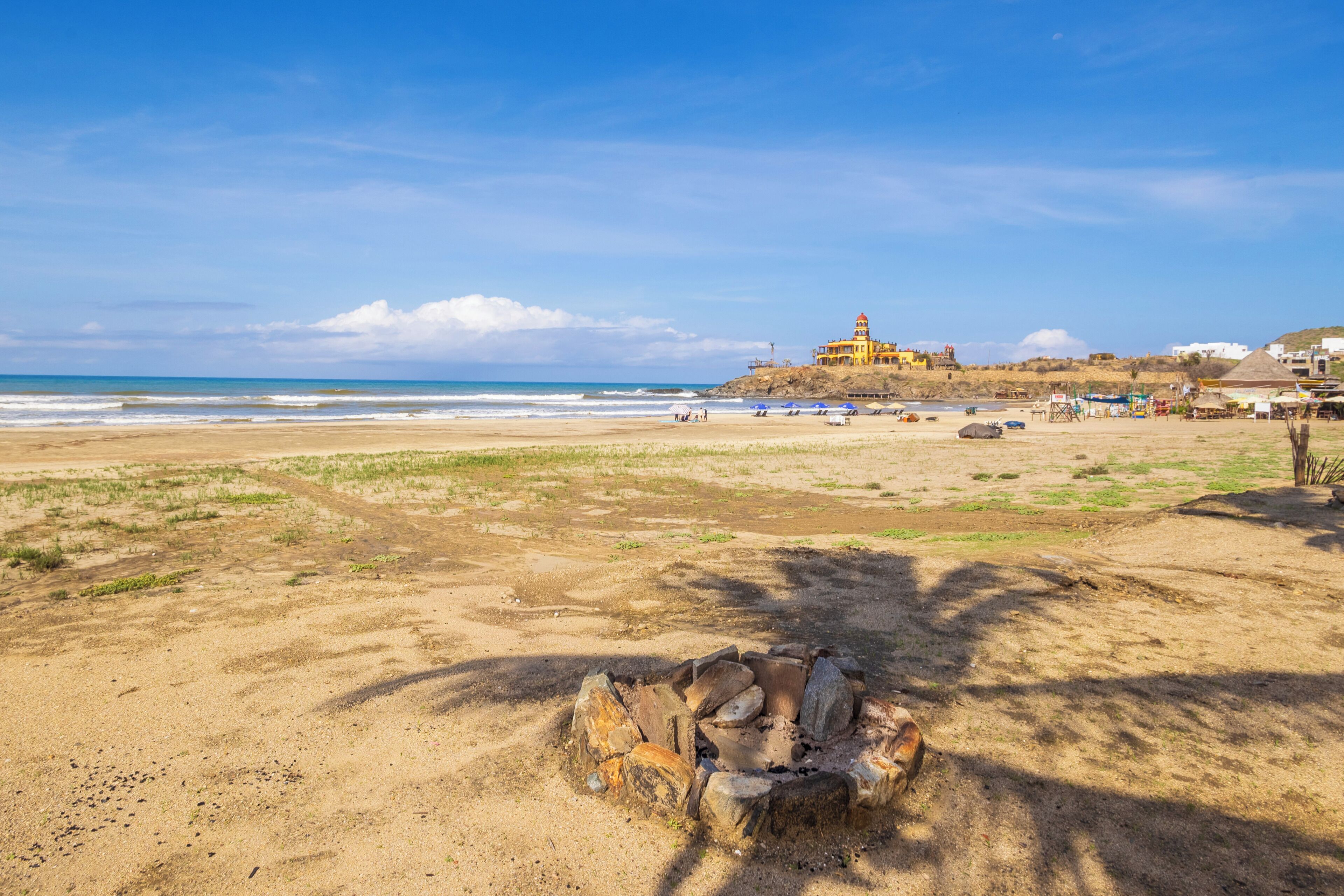 Uma praia com um edifício amarelo imponente numa colina, uma área rochosa ao primeiro plano e um céu azul límpido.