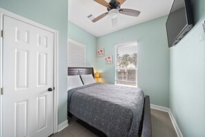 Quarto com cama, ventoinha no teto, televisão presa na parede e janela com persianas.