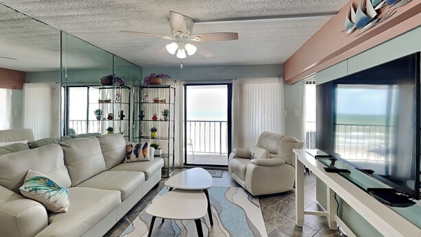 Uma sala de estar moderna com sofá, poltronas, mesa de centro e televisão. Há uma varanda com vista para o oceano.