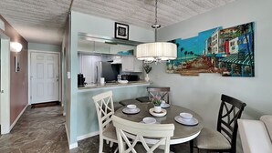 Uma sala de jantar com uma mesa redonda, cadeiras e um lustre. Uma cozinha aparece ao fundo, com armários e uma geladeira.