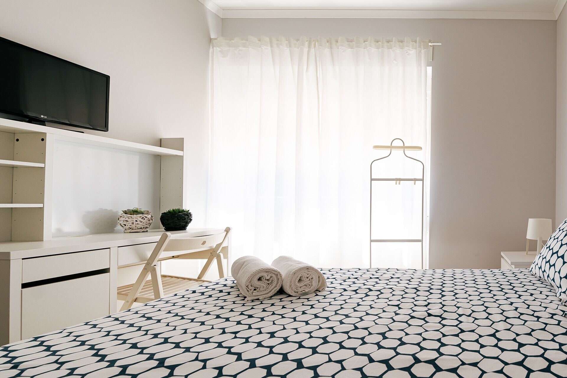 Quarto com cama, escrivaninha, cadeira, televisão, janela com cortinas e um cabideiro.
