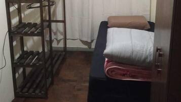 Quarto pequeno com piso de madeira, cama com almofadas, estante de metal e janela com cortinas.