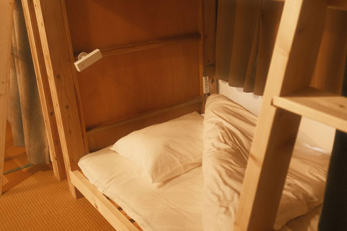 Uma cama beliche de madeira com colchão e almofadas, uma porta de madeira e uma tomada elétrica visível.