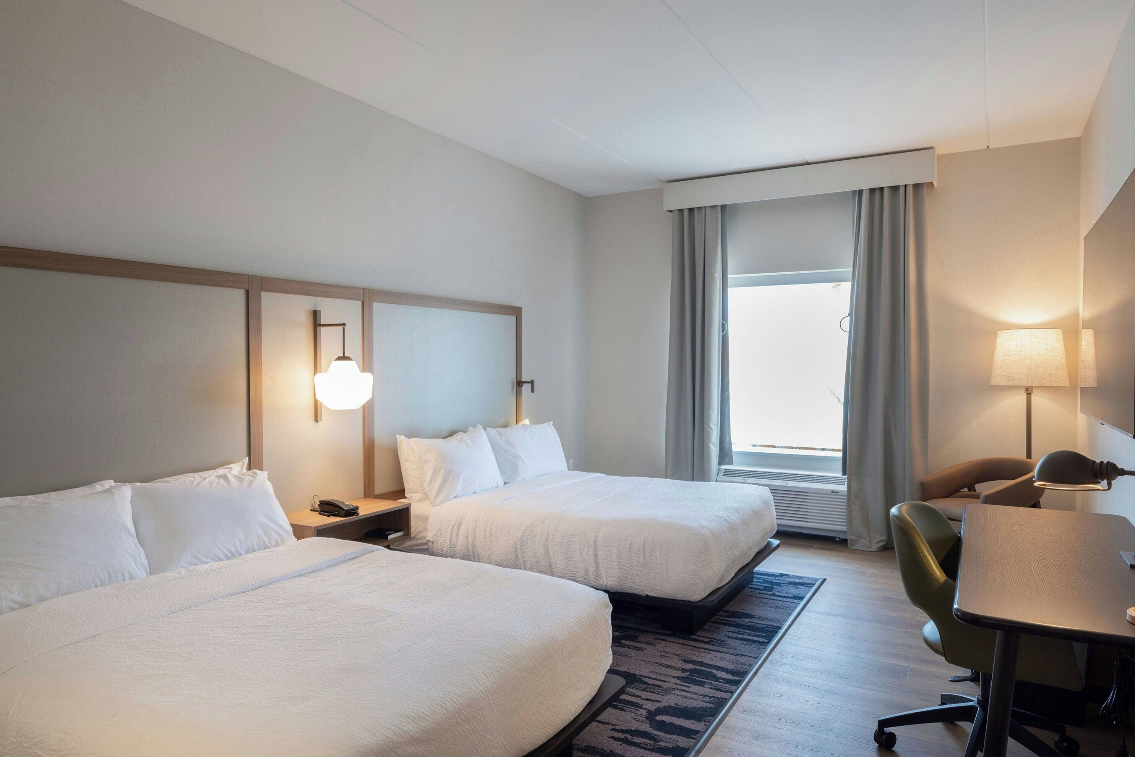 Quarto de hotel com duas camas, uma escrivaninha, uma cadeira e uma janela grande com cortinas.