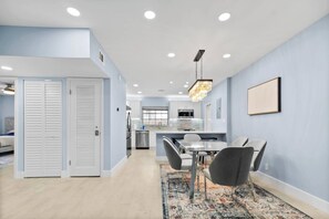 Uma cozinha moderna com uma ilha central, um espaço de refeições com mesa e cadeiras, e um corredor que dá para um quarto.
