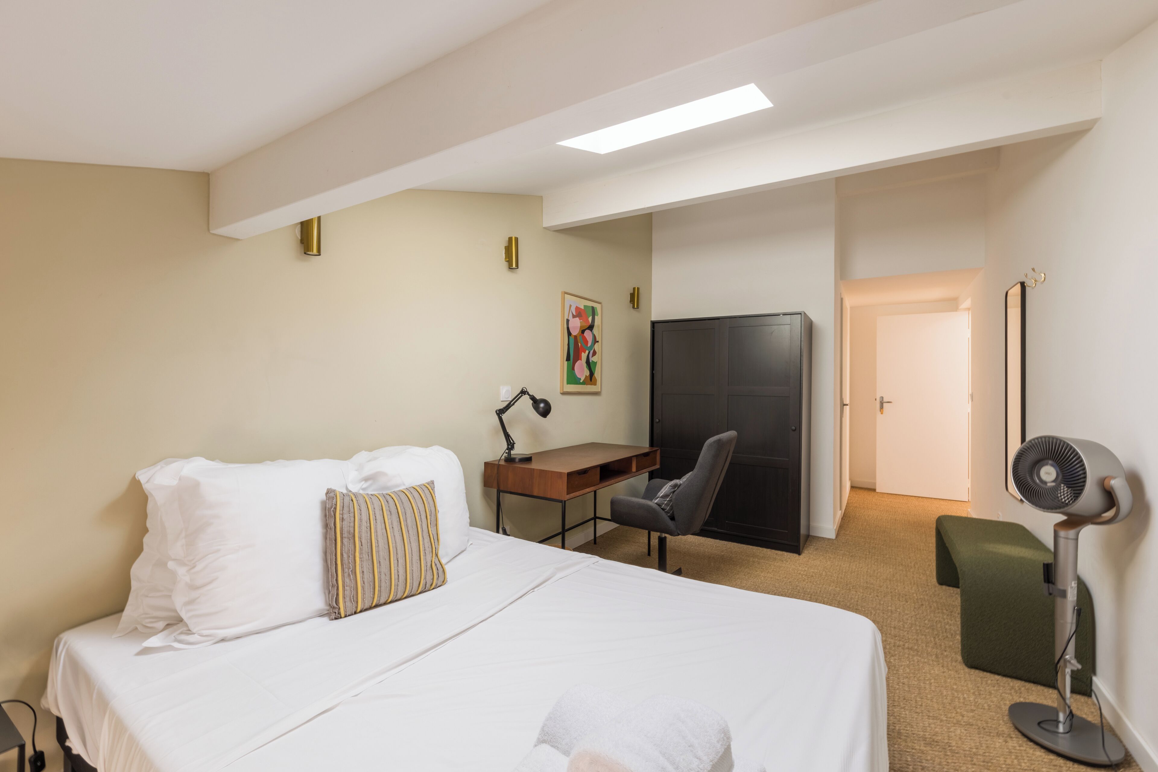 Quarto de hotel com cama, escrivaninha, cadeira, guarda-roupa e ventoinha.