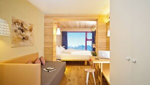 Quarto de hotel com cama, escrivaninha com laptop, um banquinho, janela com vista para as montanhas e um quadro na parede.