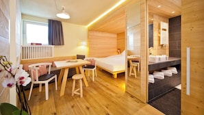 Um quarto acolhedor com piso de madeira, uma cama, uma escrivaninha, uma cadeira, um banquinho pequeno e um banheiro com pia e porta-toalhas.