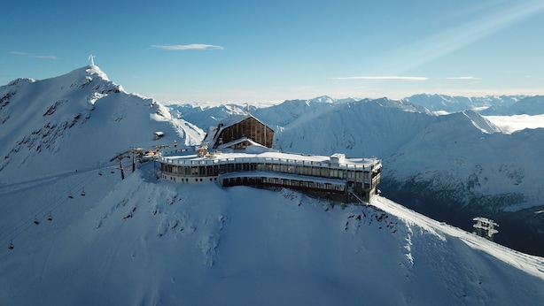 Uma pousada nos Alpes coberta de neve, com um teleférico, rodeada por picos nevados e céu limpo.