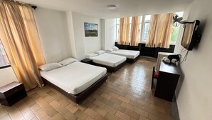Quarto de hotel com três camas, uma escrivaninha pequena, televisão e janela com cortinas.