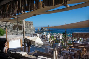 Um restaurante à beira-mar com mesas ao ar livre, vista para o oceano e um penhasco rochoso ao fundo.