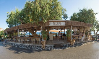 Um restaurante à beira-mar chamado "PARAISO", com mesas ao ar livre, um espaço pavimentado em pedra e vista para o mar.