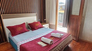 Quarto de hotel com parede de ripas de madeira, cama com roupa de cama e almofadas vermelhas, janela com cortinas e chão de madeira.