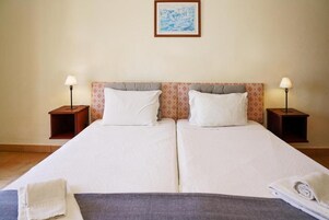 Uma cama de casal com cabeceira estampada, duas mesas de cabeceira com abajures e um quadro na parede.