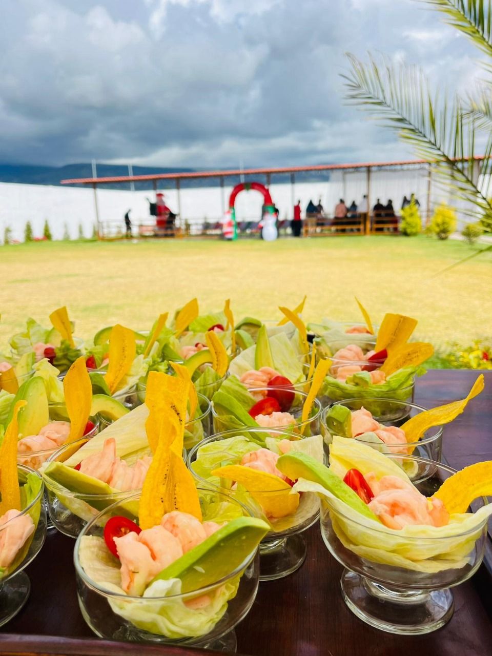 Uma mesa com petiscos de frutos do mar disposta ao ar livre, com vista para uma área de estar coberta e um céu nublado ao fundo.