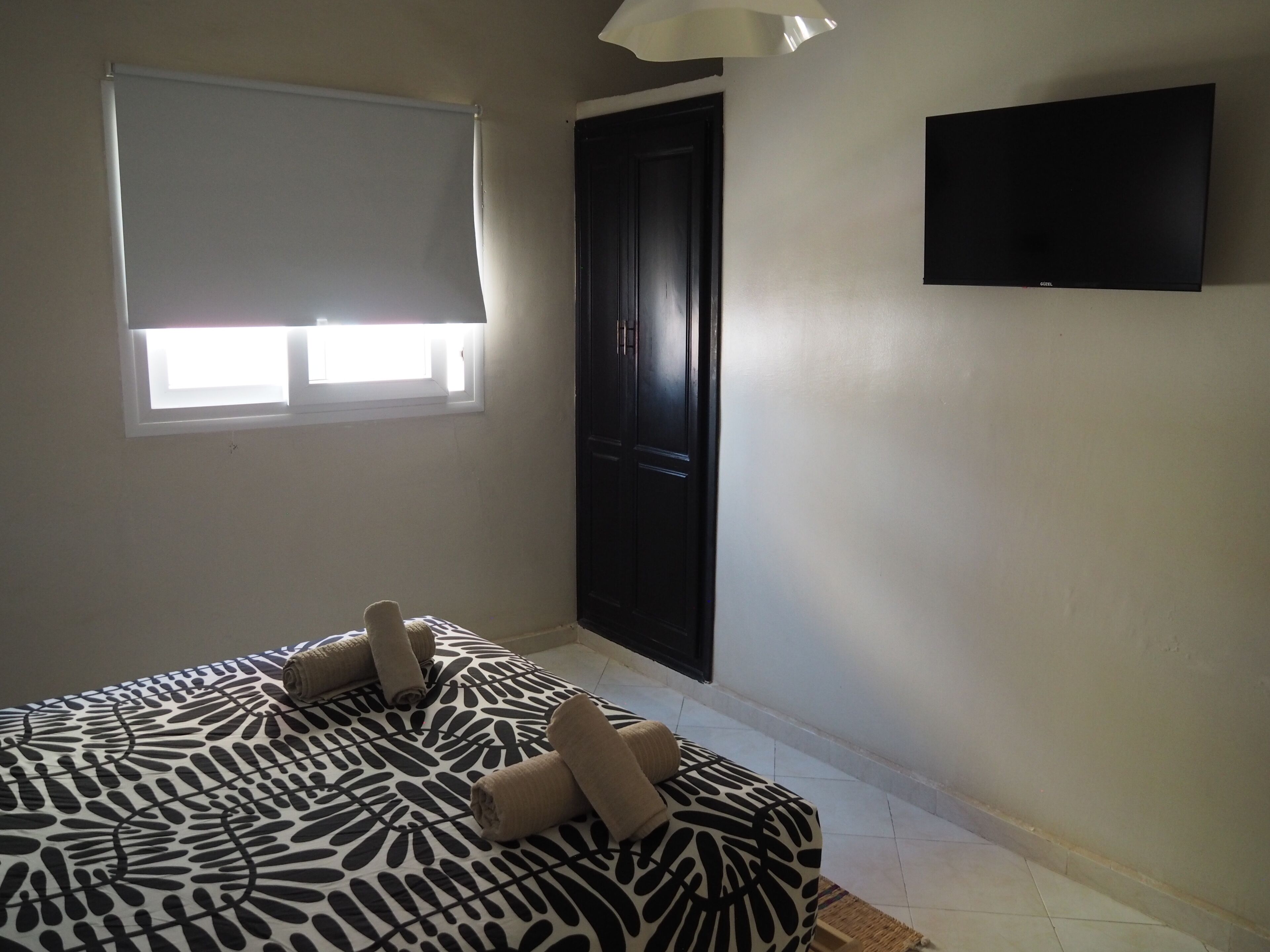 Quarto com cama, janela com persianas, porta e televisão na parede.