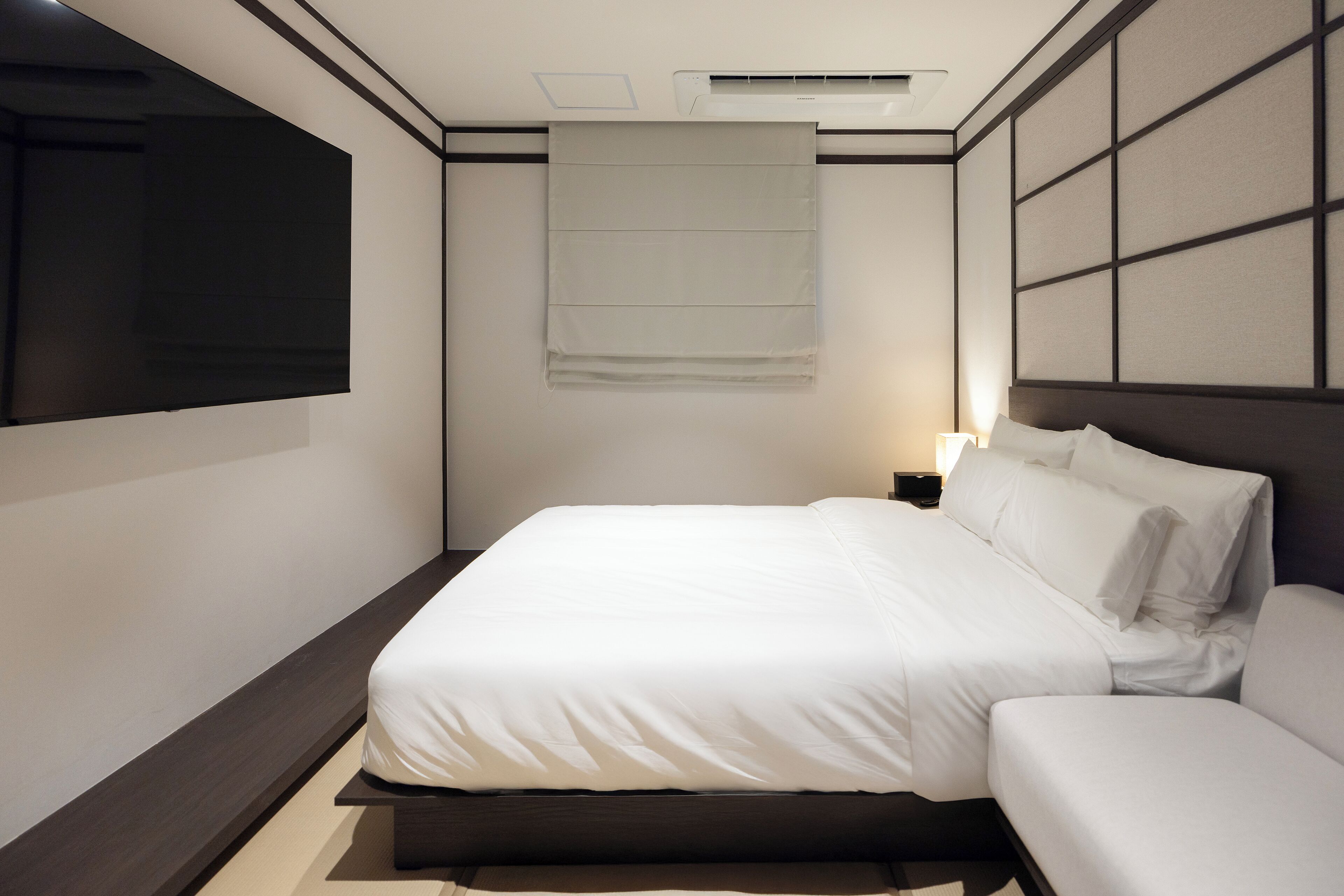 Quarto de hotel moderno com uma cama grande, uma TV de tela plana na parede e uma mesa de cabeceira com abajur.