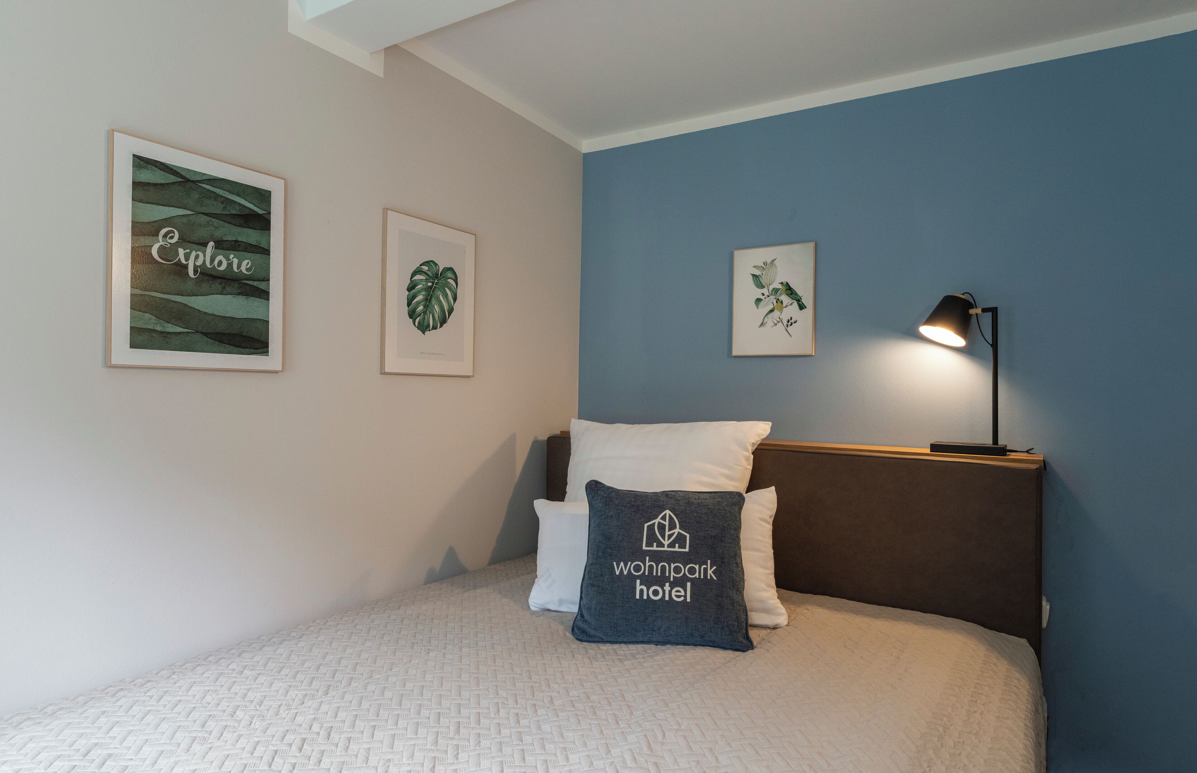 Quarto com cama, travesseiros, mesa de cabeceira, abajur e quadros na parede.