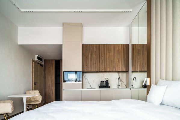 Quarto de hotel moderno com uma cama grande, um espaço de estar pequeno, uma kitchenette e um espelho amplo.