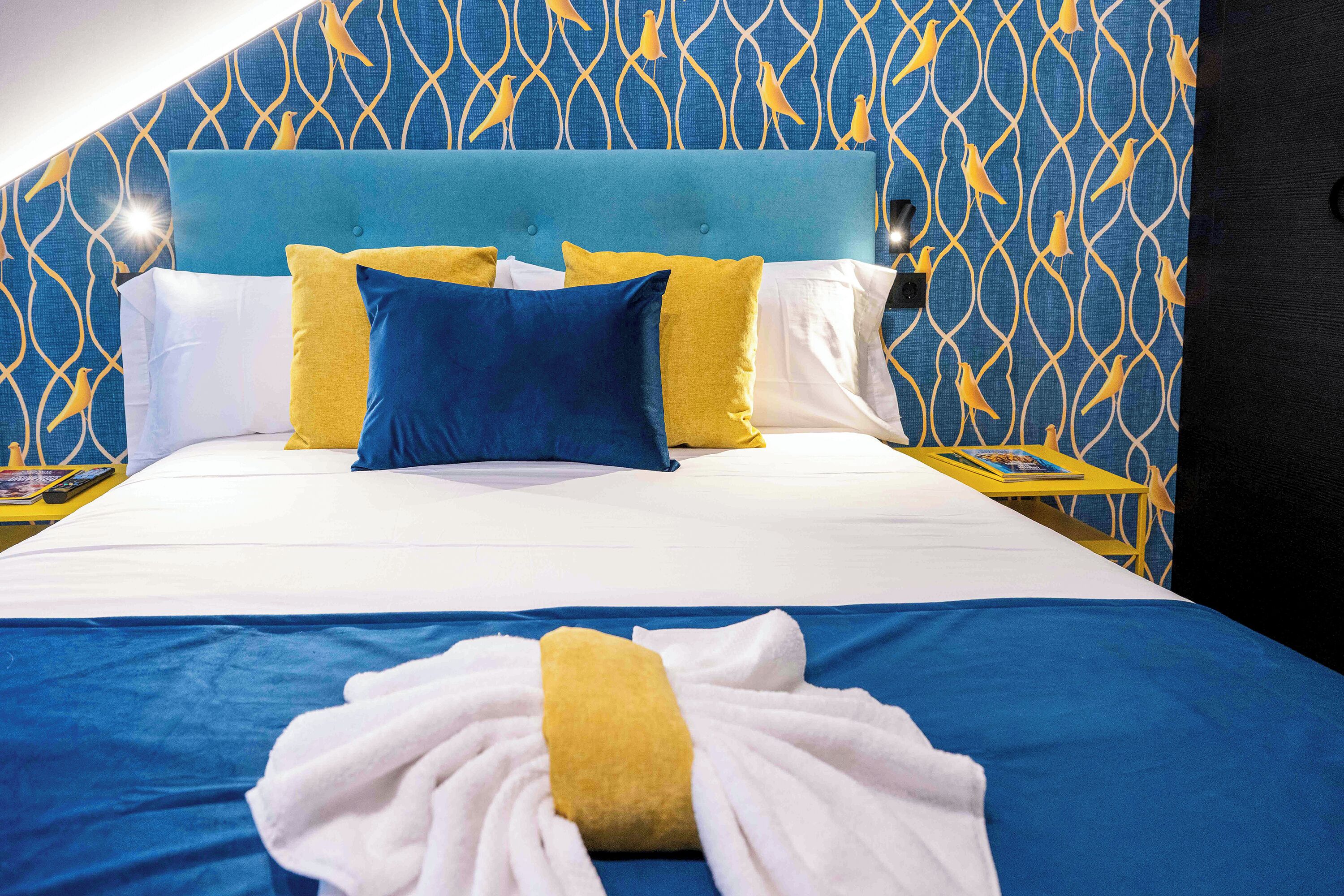 Quarto de hotel com parede estampada em azul e amarelo, cabeceira azul e cama bem arrumada, com toalha e uma toalha enrolada sobre a cama.