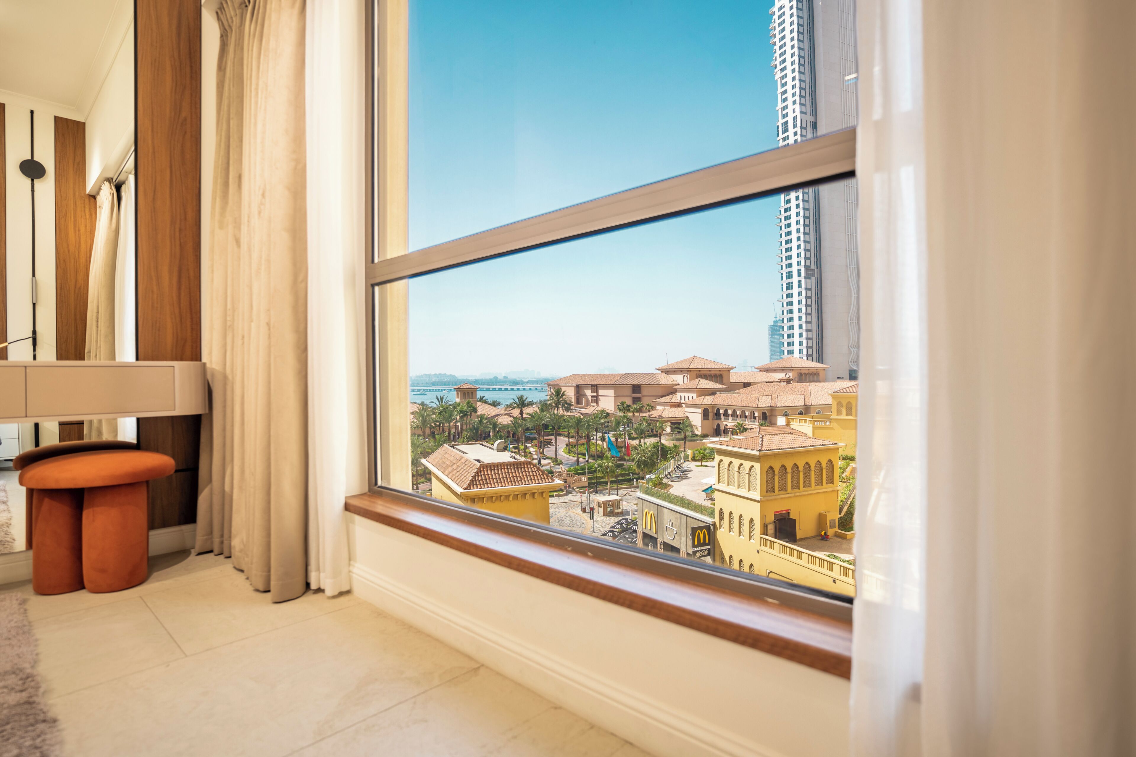 Quarto com uma janela grande, piso de madeira, um banquinho laranja pequeno e vista para um horizonte urbano com edifícios e palmeiras.