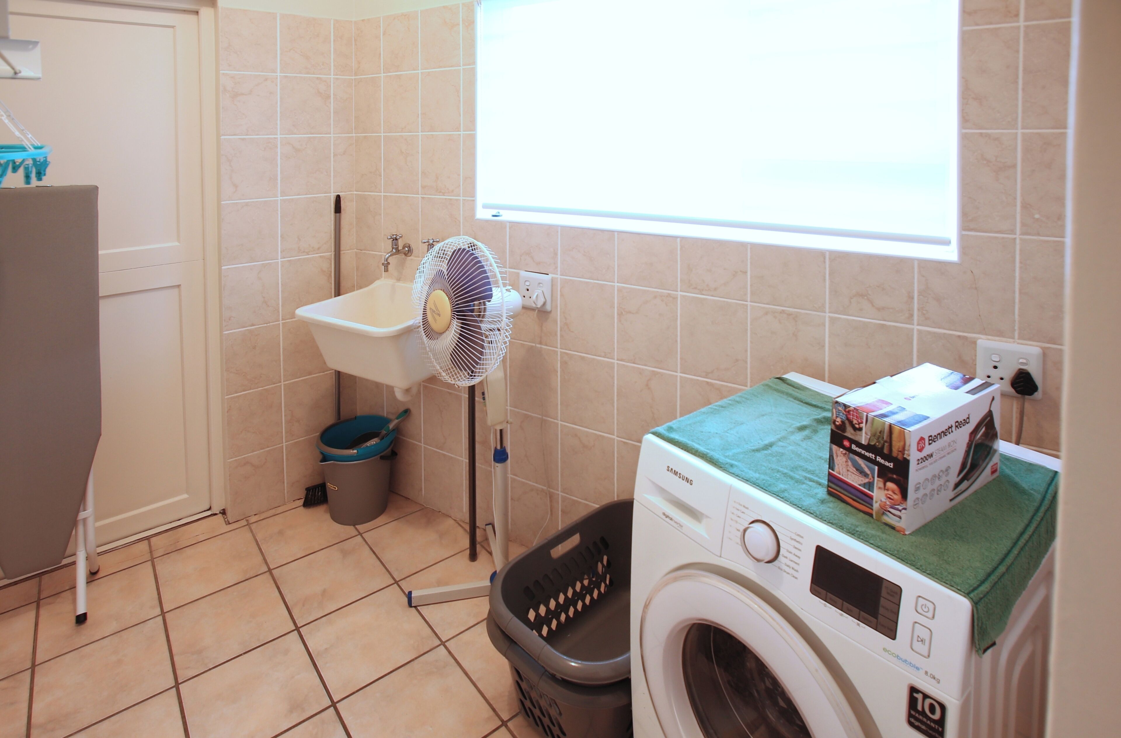 Uma lavanderia com máquina de lavar, pia, ventilador e janela.