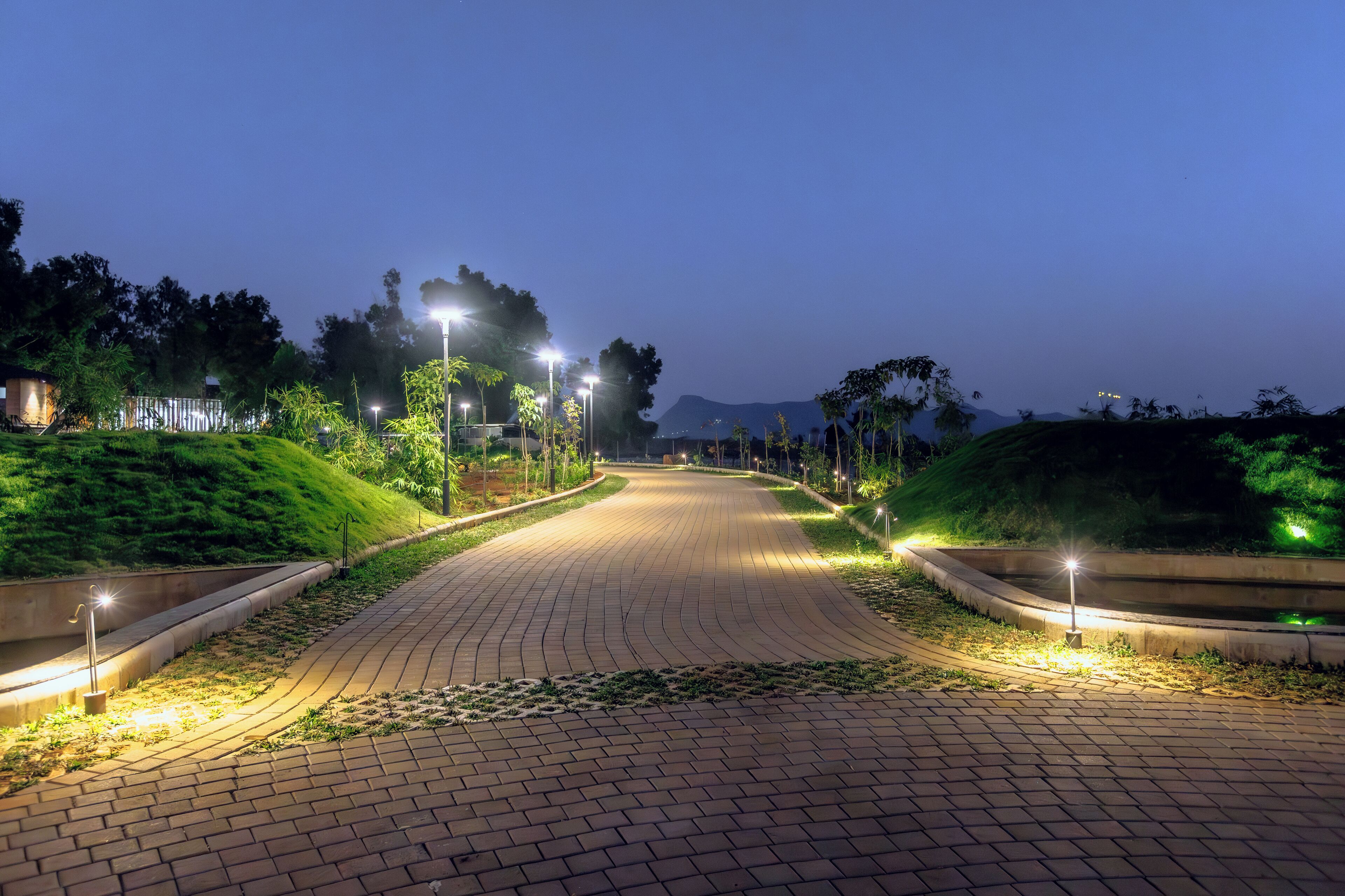 Um caminho pavimentado, ladeado por áreas gramadas e árvores, conduz a uma colina distante, com postes de iluminação.