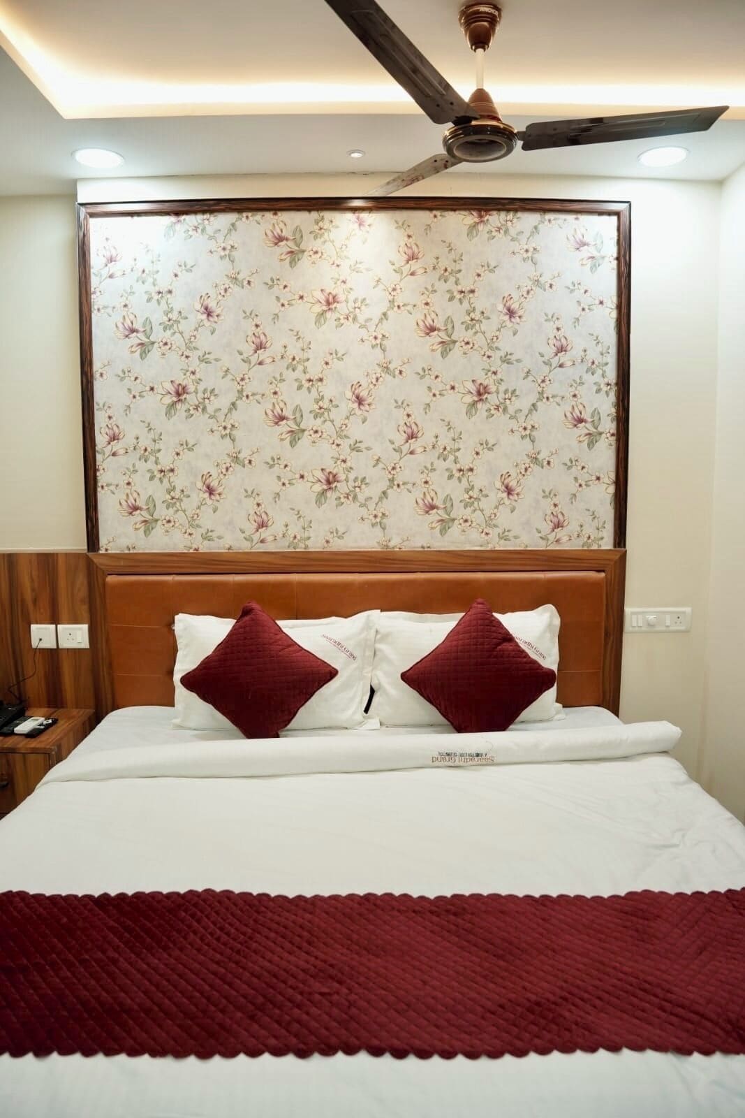 Quarto de hotel com cabeceira de papel de parede floral, cabeceira de madeira, duas almofadas vermelhas e cama com roupa de cama branca.
