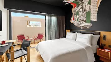 Quarto de hotel com uma cama grande, uma escrivaninha com cadeira, uma mesinha e vista para o exterior através de uma porta deslizante.