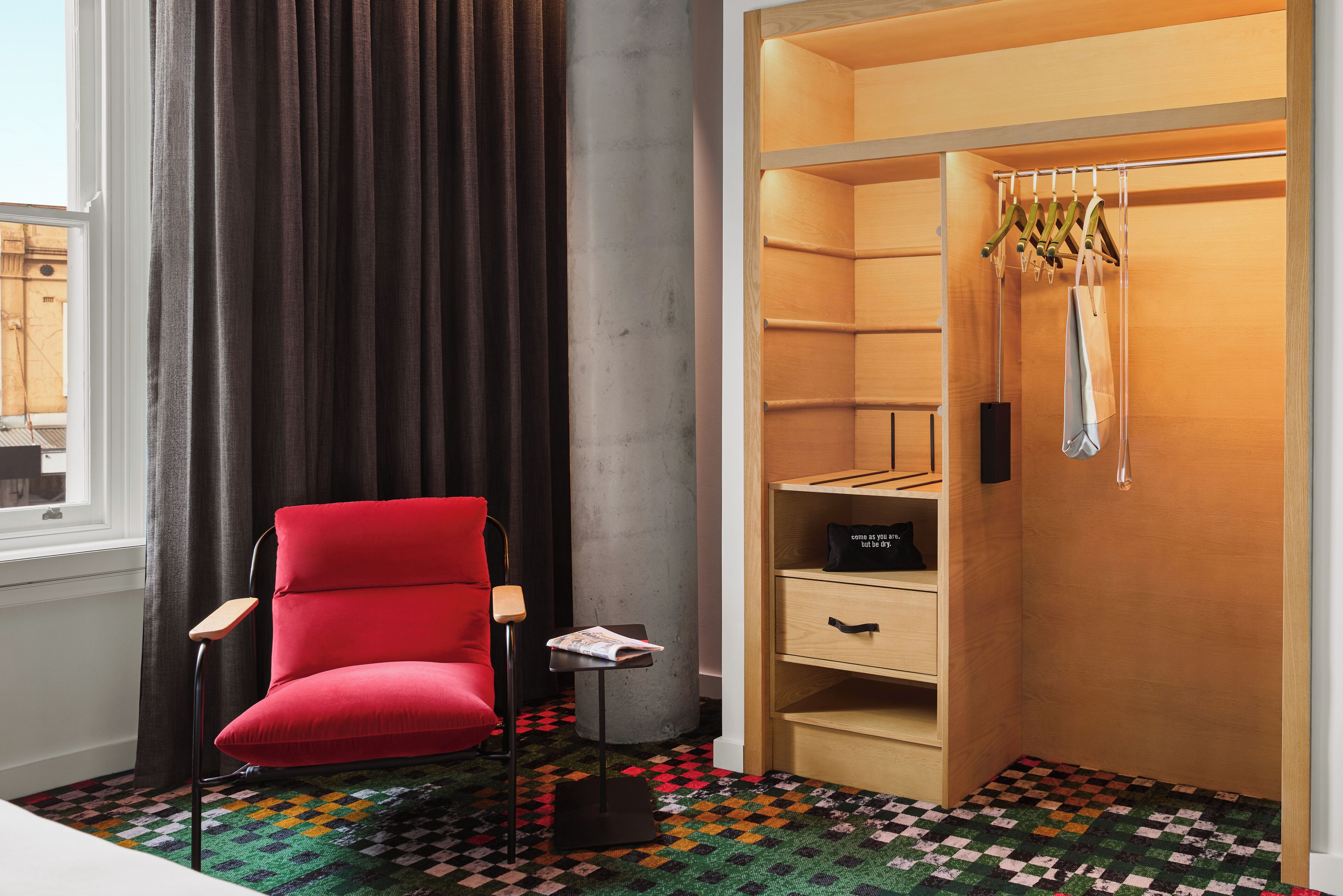 Quarto com uma cadeira vermelha, um guarda-roupa de madeira com espaço para pendurar roupas e um tapete colorido estampado.