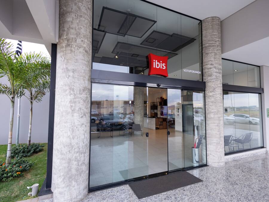 Um edifício moderno com entrada de vidro, um letreiro vermelho chamativo da marca 'ibis' e uma área de estacionamento para carros visível na parte externa.